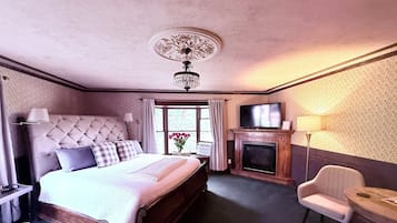 Quarto de hotel com uma cama grande, televisão, lareira, uma cadeira e janela com cortinas.
