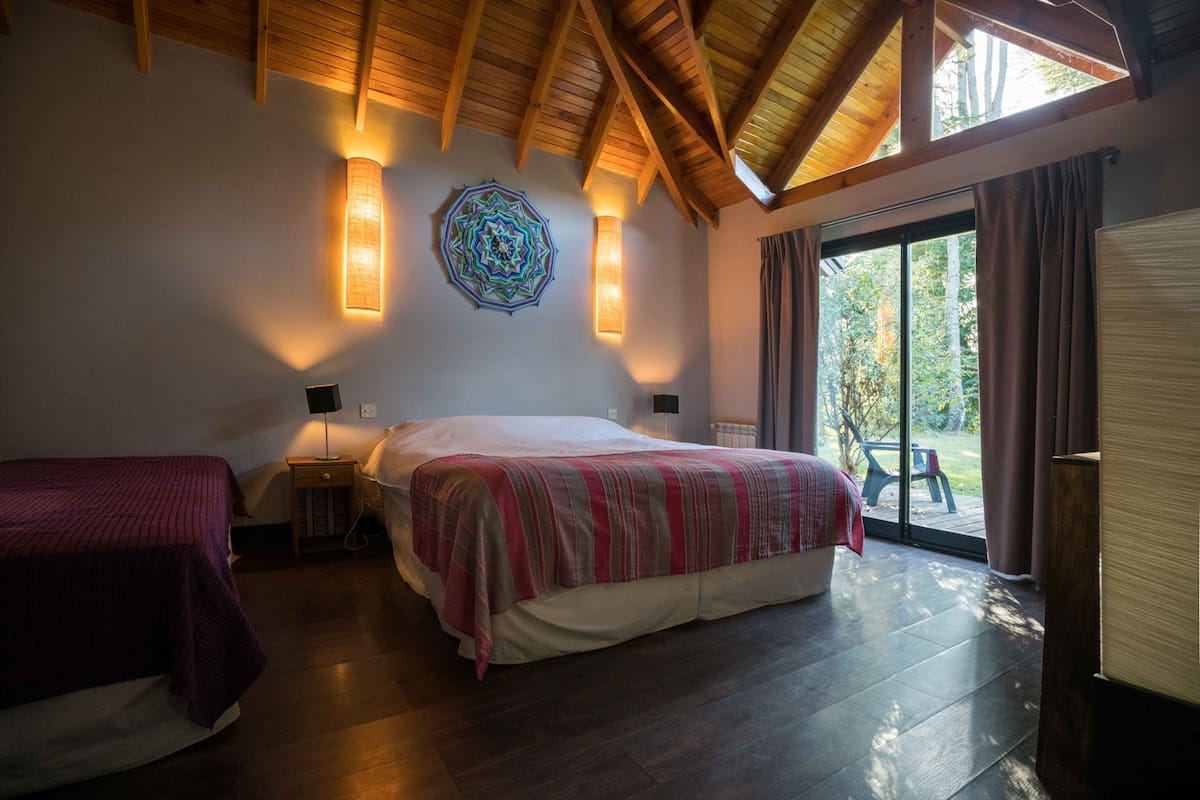 Quarto com uma cama grande, uma cama menor, uma mesa de cabeceira, um abajur, uma obra de arte na parede e uma porta de vidro deslizante para uma área externa.