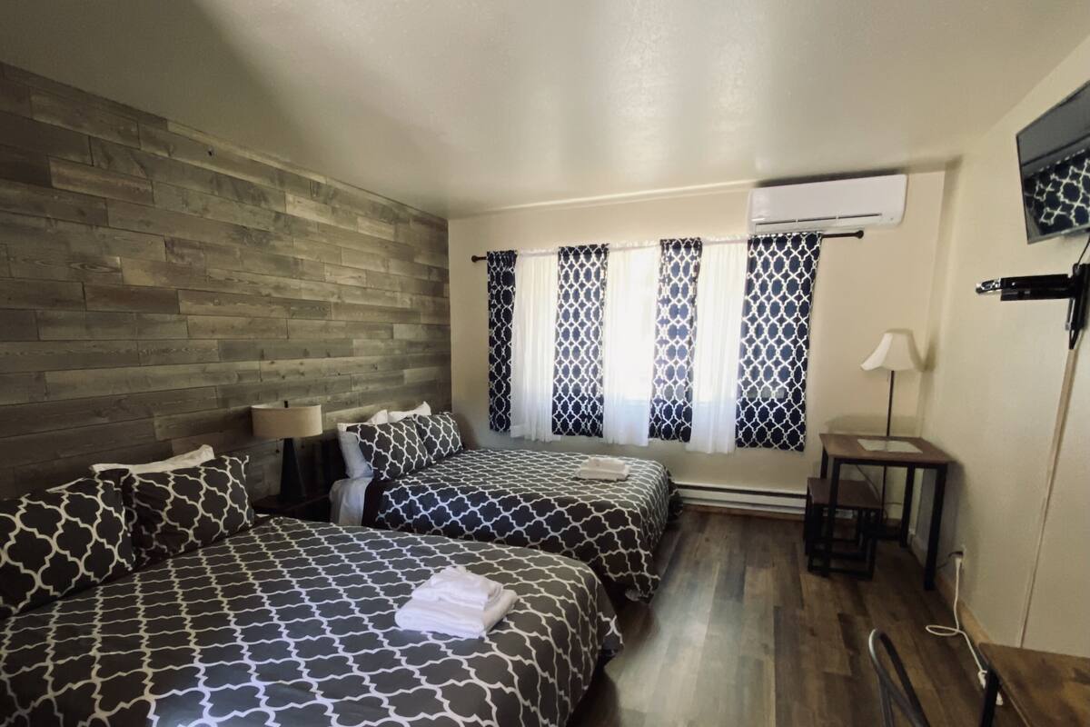 Quarto com duas camas, parede de madeira, janela com cortinas estampadas, escrivaninha e uma cadeira.