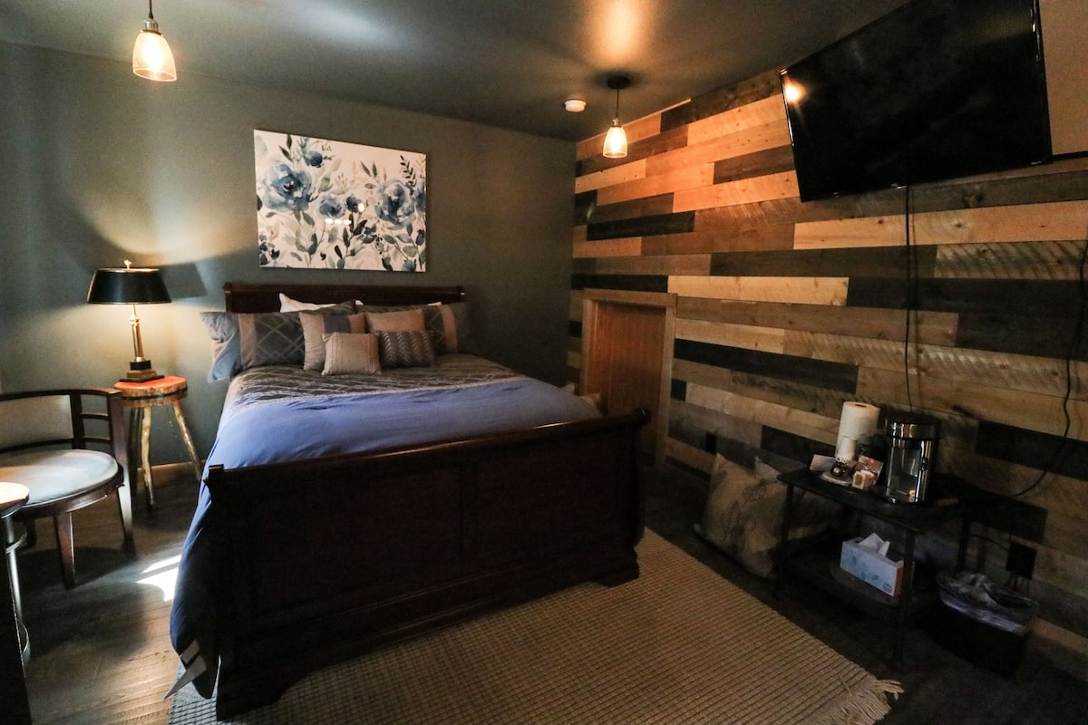 Quarto com cabeceira de madeira, cama com roupa de cama azul, mesa de cabeceira com abajur, um quadro na parede e TV na parede.