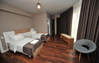 Quarto de hotel moderno com duas camas, um banco, uma cadeira e uma janela grande com cortinas.