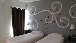 Quarto com duas camas de solteiro, uma mesa de cabeceira com abajur, uma janela com cortinas e uma parede com padrão de folhas.