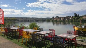 Mesas e cadeiras ao ar livre perto de um lago, com vista para edifícios e uma ponte ao fundo.