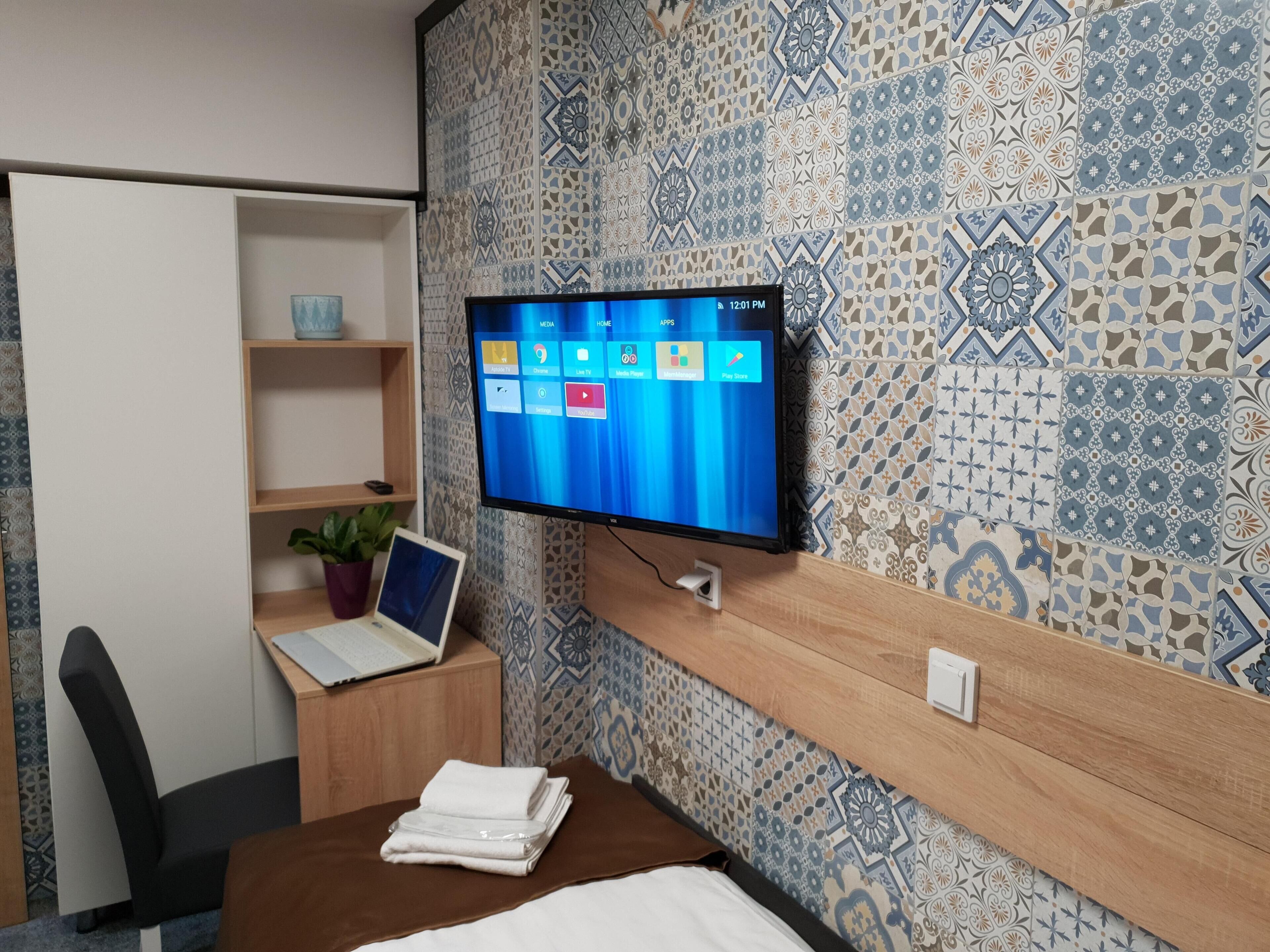 Quarto de hotel com televisão de tela plana na parede, uma escrivaninha de madeira com um laptop, uma cadeira e uma cama com cabeceira marrom.