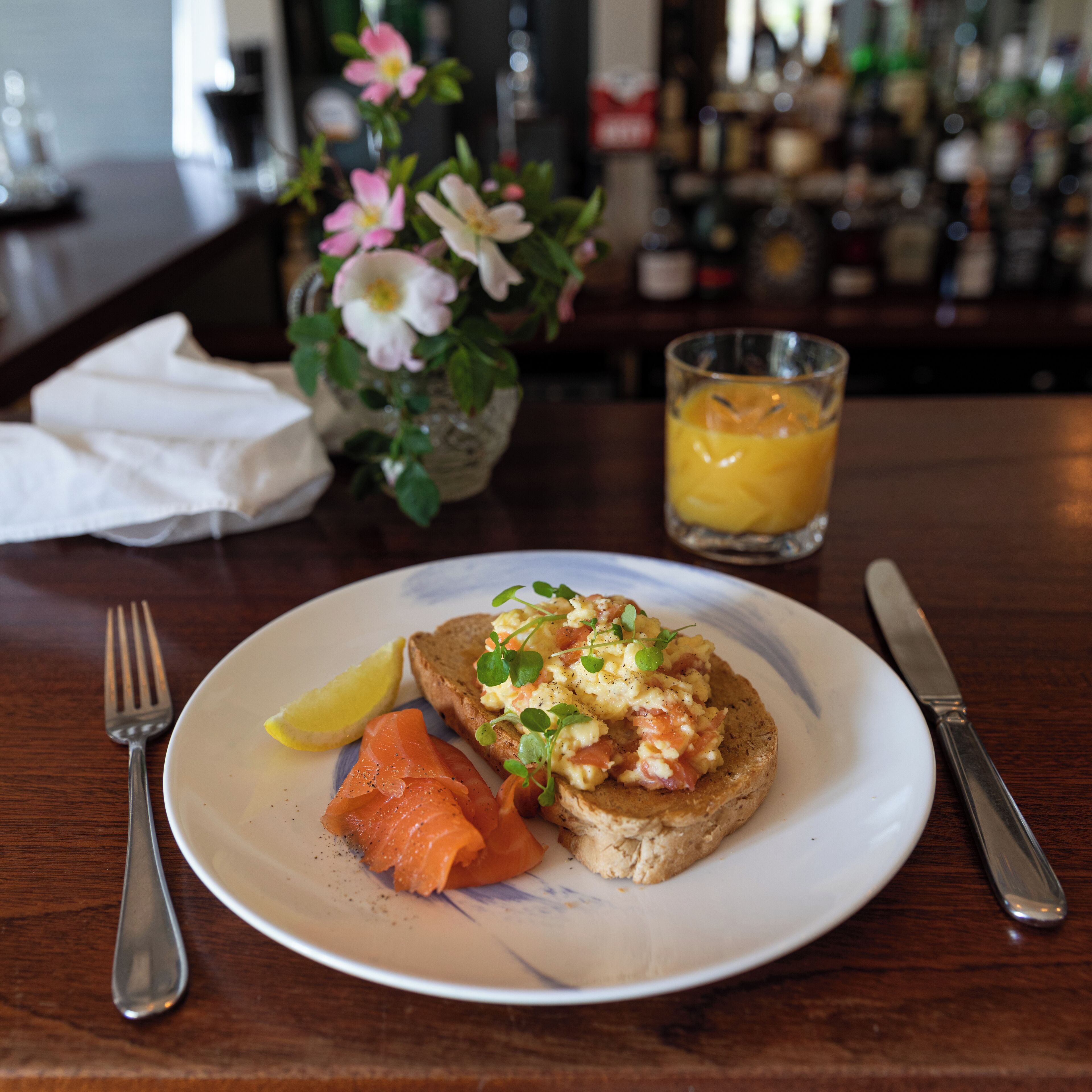 Prato de café da manhã com torradas, salmão e ovos mexidos, um copo de suco de laranja e um vaso com flores sobre uma mesa de madeira.