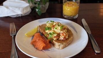 Prato de café da manhã com torradas, salmão e ovos mexidos, um copo de suco de laranja e um vaso com flores sobre uma mesa de madeira.