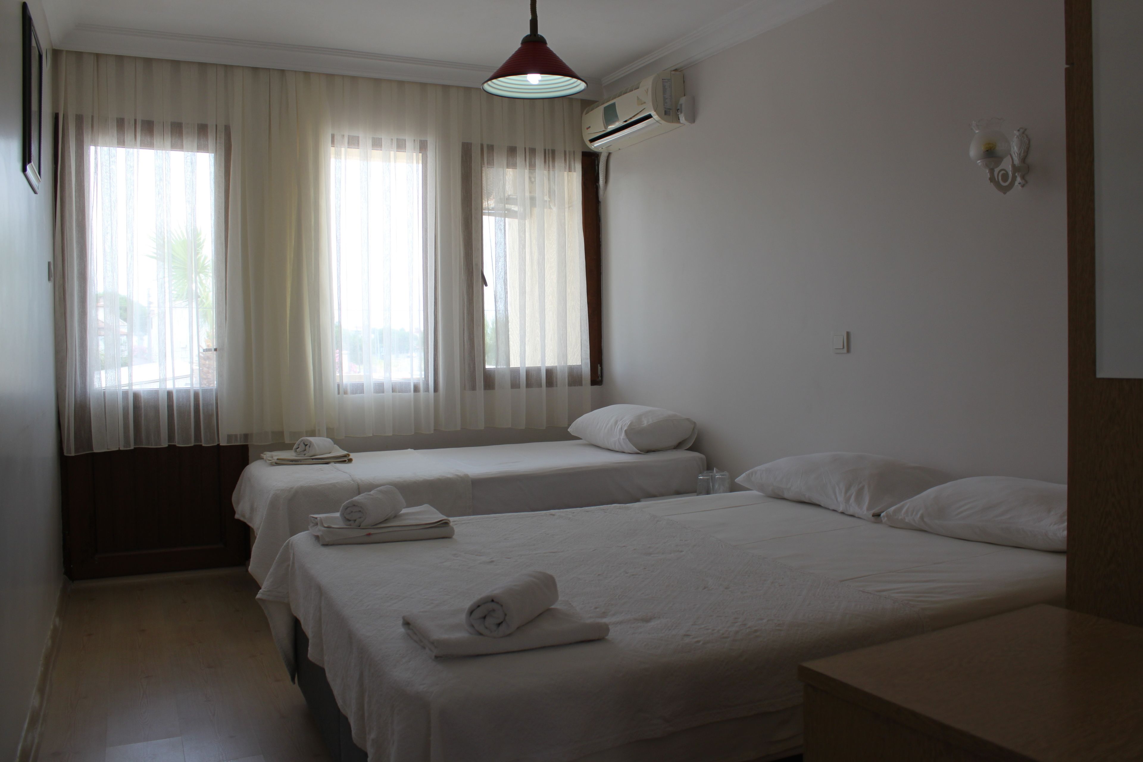Quarto de hotel com duas camas, uma mesa de cabeceira de madeira, uma luminária pendente vermelha e um aparelho de ar condicionado na parede.