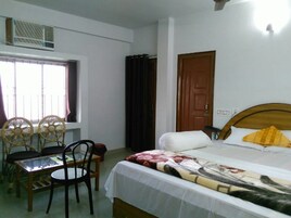 Quarto com cama, cabeceira de madeira, janela com cortinas, mesa com cadeiras e aparelho de ar condicionado.