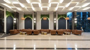 Um hall espaçoso com design moderno, apresentando uma área de estar com sofás e mesas redondas, teto decorativo com iluminação integrada e uma grande janela com vista para o exterior.