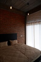 Cama com cabeceira em frente a uma parede de tijolos, janela com cortinas e duas lâmpadas penduradas.
