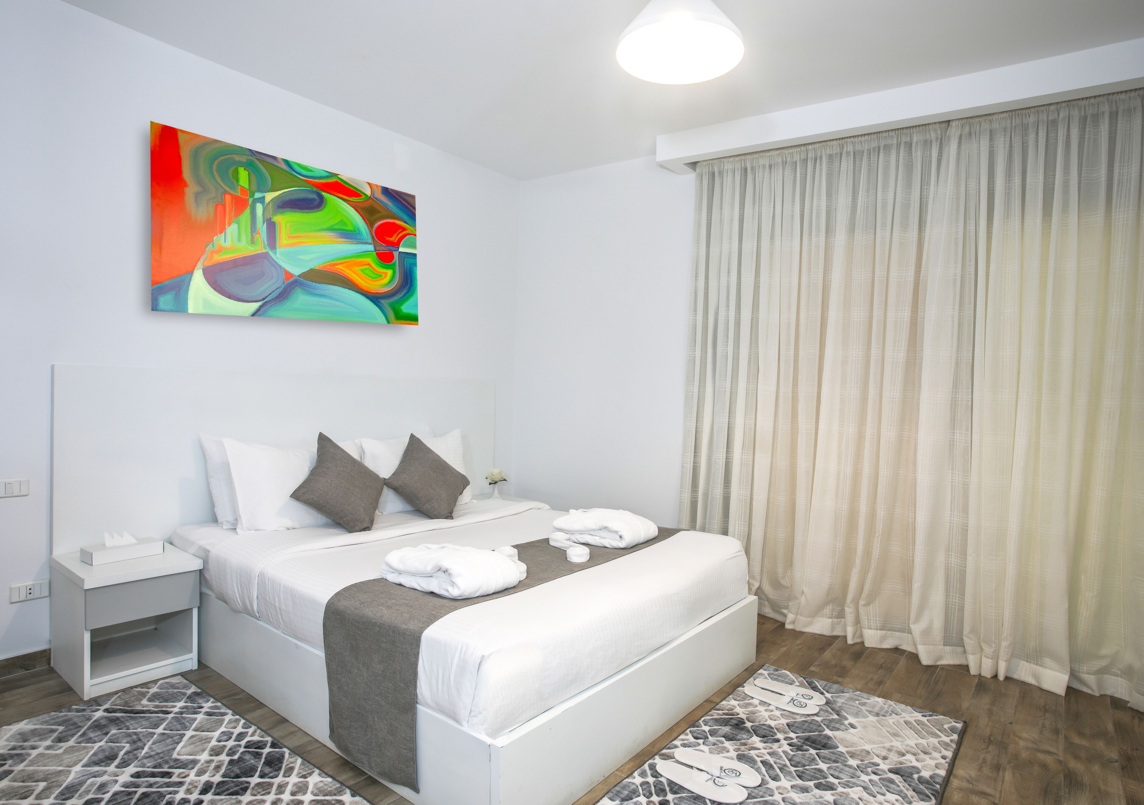 Quarto de hotel moderno com uma cama grande, mesa de cabeceira, luminária e um quadro abstrato colorido, além de uma janela ampla com cortinas leves.