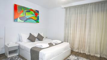 Quarto de hotel moderno com uma cama grande, mesa de cabeceira, luminária e um quadro abstrato colorido, além de uma janela ampla com cortinas leves.