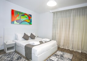 Quarto de hotel moderno com uma cama grande, mesa de cabeceira, luminária e um quadro abstrato colorido, além de uma janela ampla com cortinas leves.