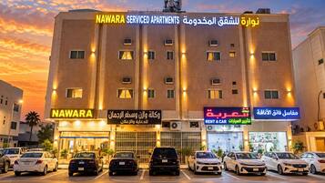Edifício com vários andares, placa com 'NAWARA SERVICED APARTMENTS', entrada com portão e vários carros estacionados em frente.