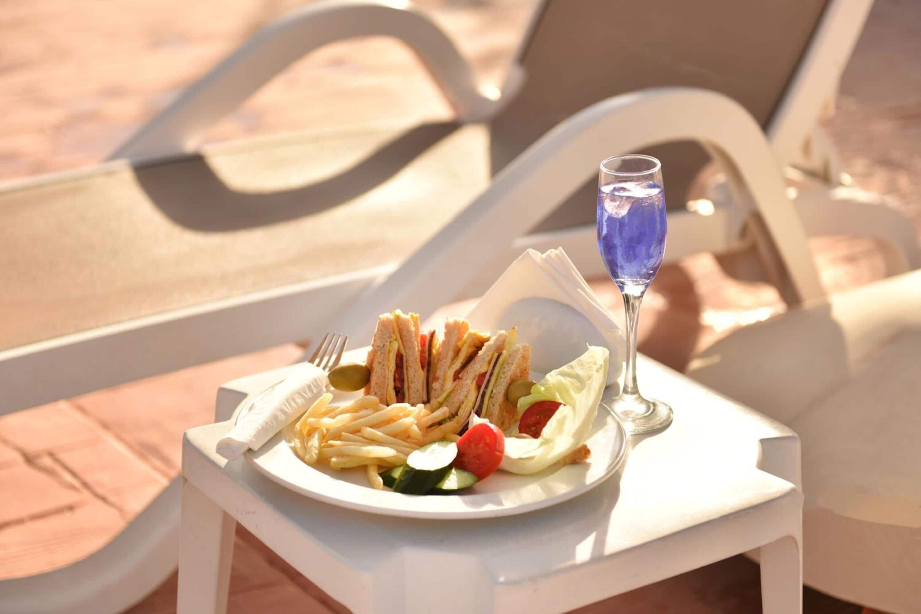 Prato de comida com sanduíche, batatas fritas e legumes em mesa branca ao ar livre.