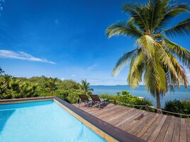 Um deck de madeira com piscina e espreguiçadeiras, com vista para a água e palmeiras.