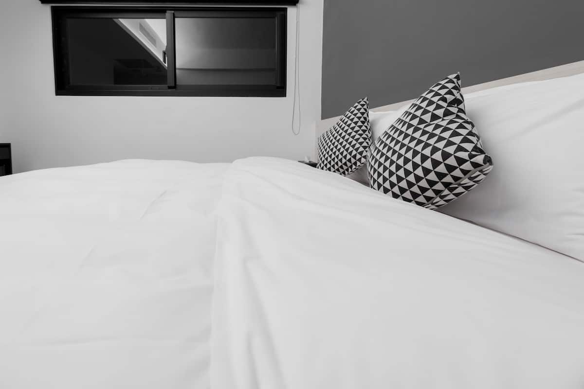 Cama bem arrumada com roupa de cama branca e almofadas estampadas em preto e branco, mesa de cabeceira preta com um objeto vermelho e uma janela com persianas.