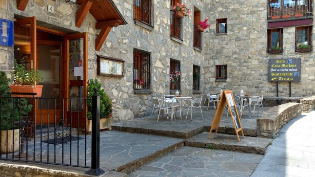 Edifício de pedra com terraço, mesas ao ar livre e uma placa indicando um restaurante.
