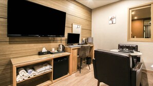Quarto de hotel com televisão de tela plana, uma escrivaninha com computador, uma cadeira, um refrigerador pequeno e uma prateleira com toalhas.
