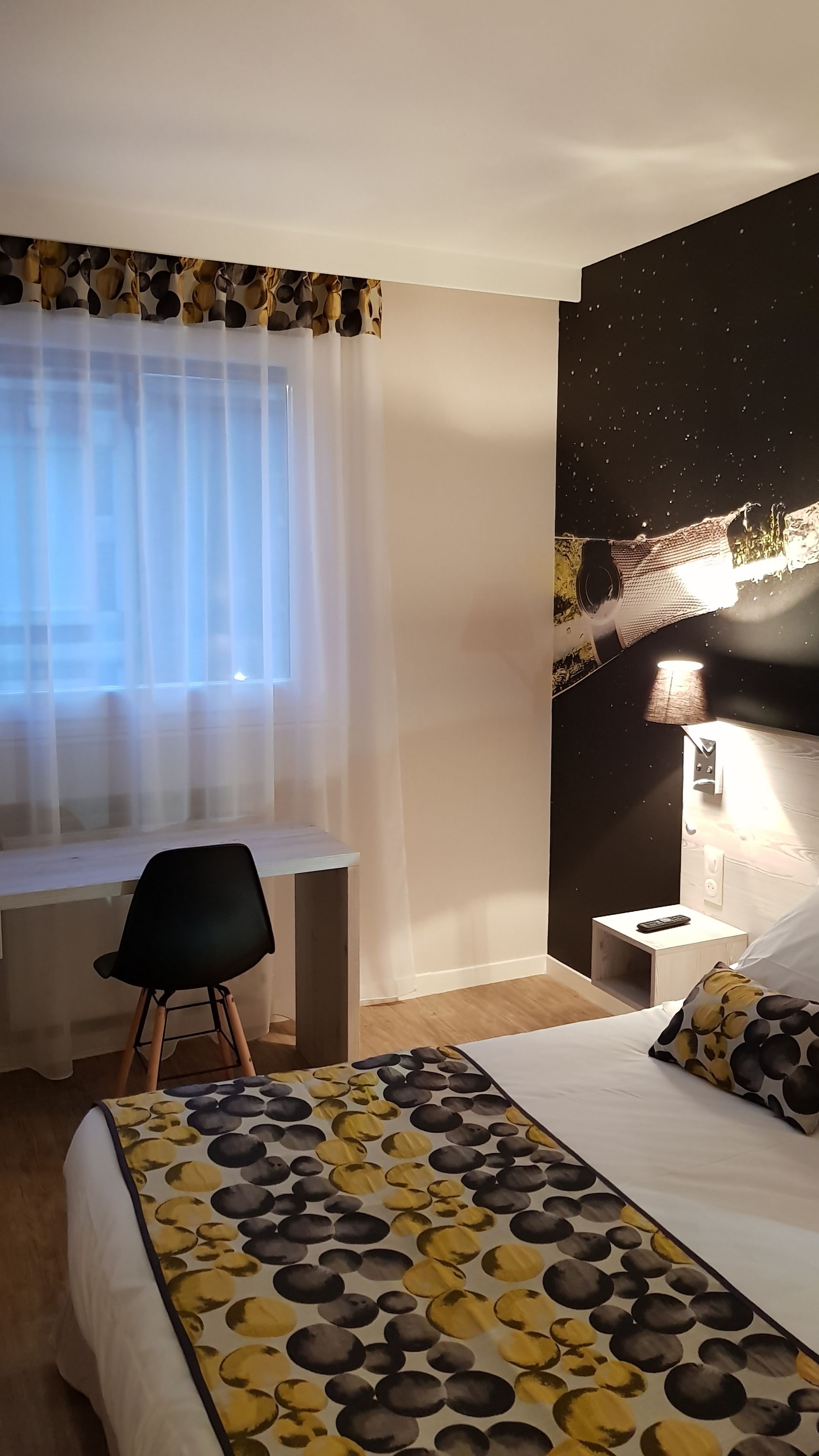 Quarto de hotel com cama, escrivaninha, cadeira, mesa de cabeceira e parede com padrão decorativo.
