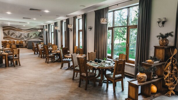 Uma ampla sala de jantar com mesas e cadeiras de madeira, janelas grandes com cortinas e luminárias fixadas na parede.