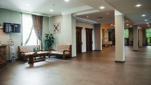 Um hall amplo com um balcão de recepção de madeira, uma área de estar confortável com sofá e poltronas, e uma televisão grande fixada na parede.