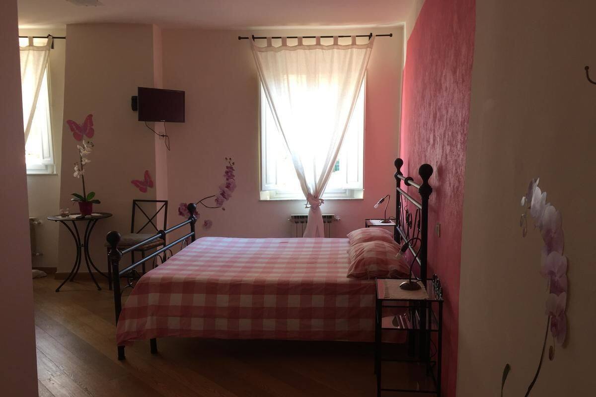 Quarto com colcha xadrez, mesa de cabeceira, uma cadeira, televisão presa à parede e janela com cortinas leves.