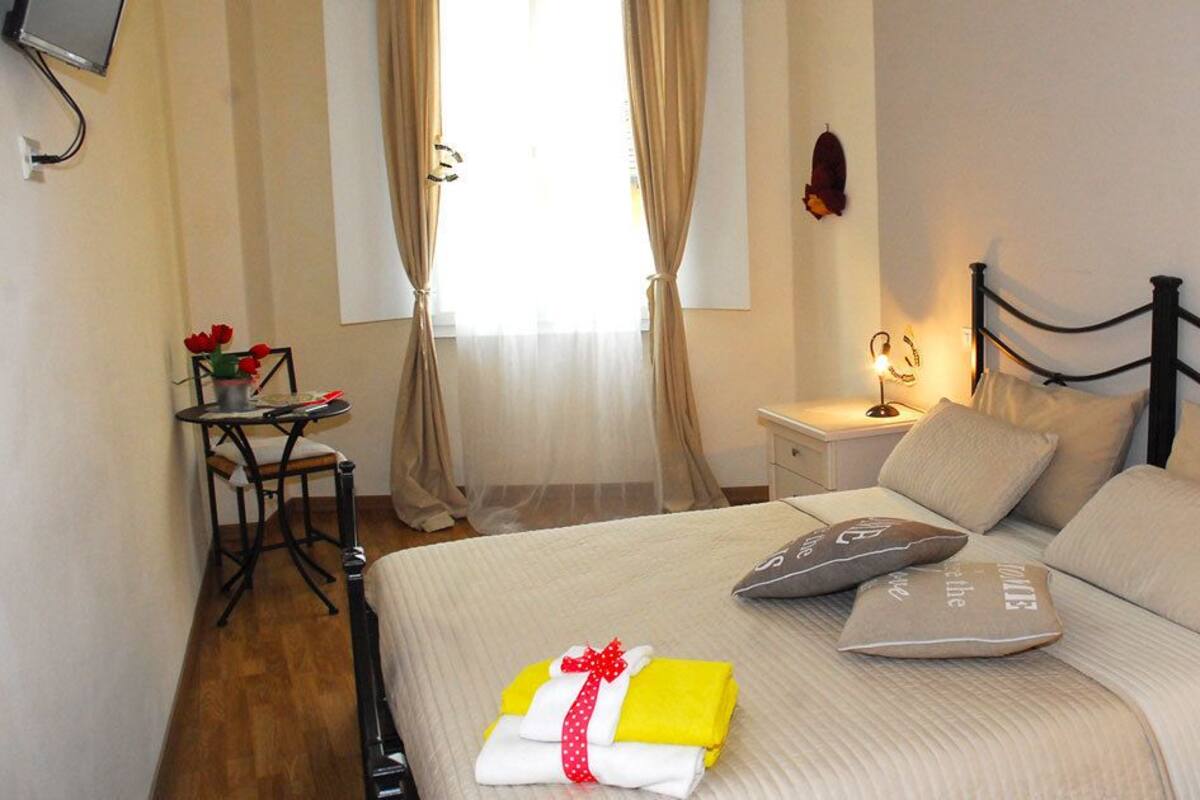 Cama bem arrumada com travesseiros e toalha dobrada, mesa de cabeceira com abajur, pequena mesa com um vaso de flores e uma janela com cortinas.