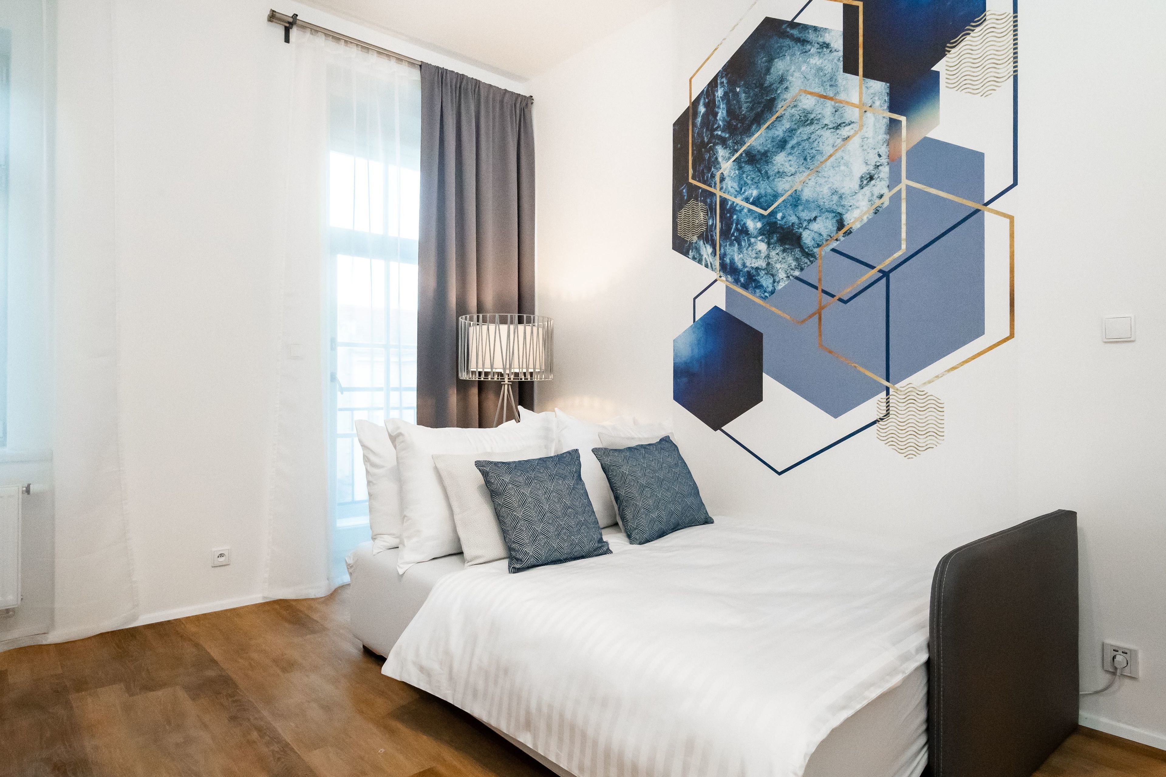Quarto moderno com cama, mesa de cabeceira, abajur e uma obra de arte geométrica na parede.