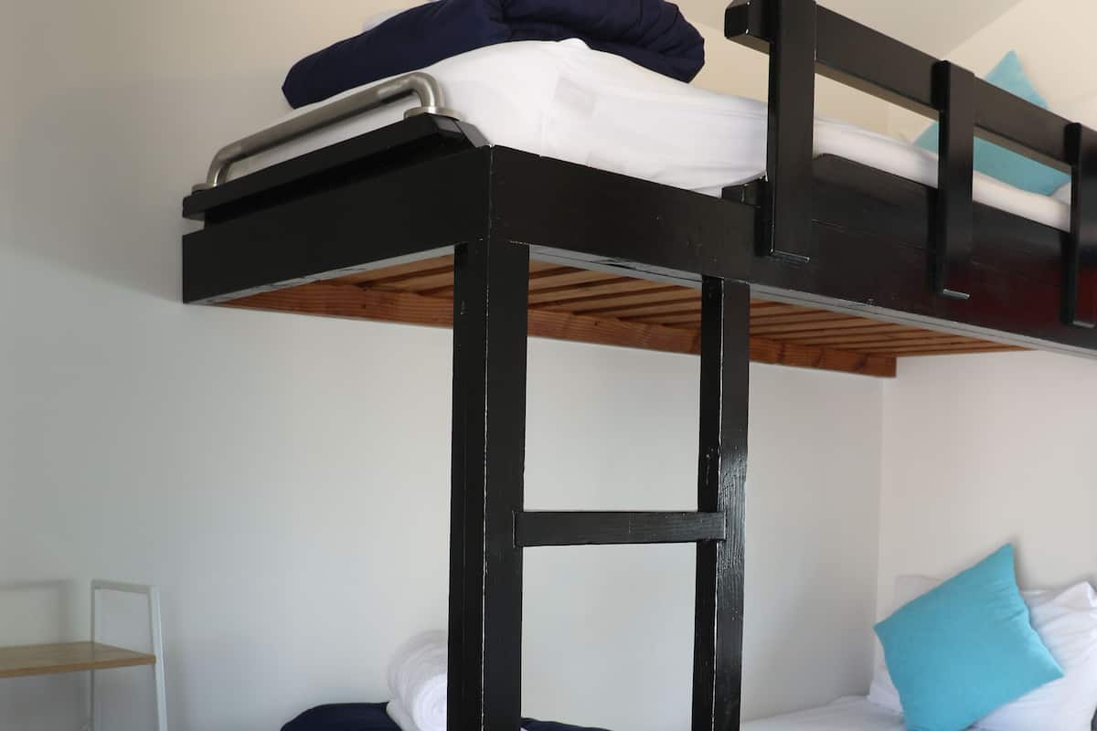 Uma cama beliche com roupa de cama branca e almofadas azuis, uma escada de madeira e uma estante branca ao lado.