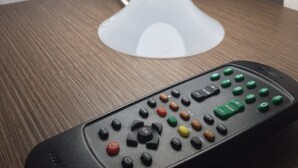 Um controle remoto sobre uma superfície de madeira, com uma luminária branca ao fundo.