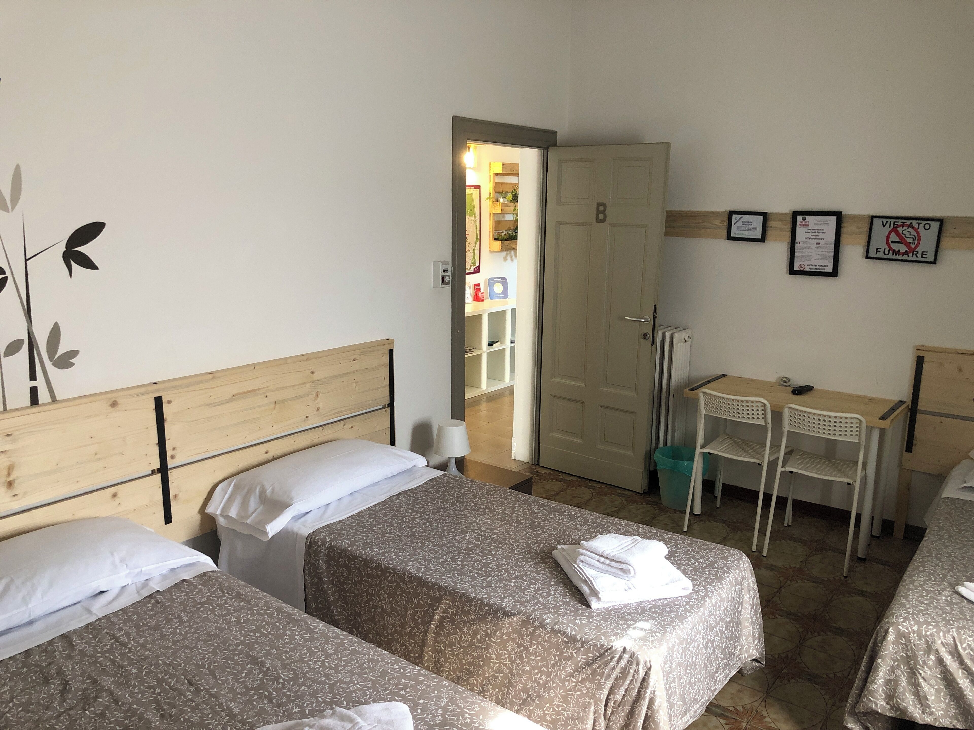 Quarto de hotel com duas camas, uma mesa de trabalho e uma cadeira. Há uma porta que dá para o banheiro e uma pequena prateleira com produtos de higiene pessoal.