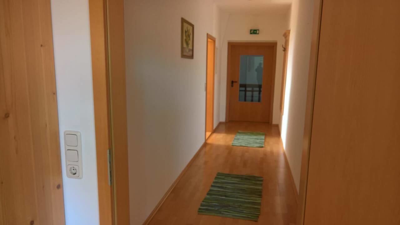 Um corredor com assoalho de madeira e tapetes verdes, que leva a uma porta com painel de vidro e sinalização de saída verde.