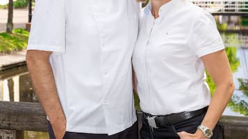 Duas pessoas ao lado de um corrimão de madeira, com um corpo de água e vegetação ao fundo.