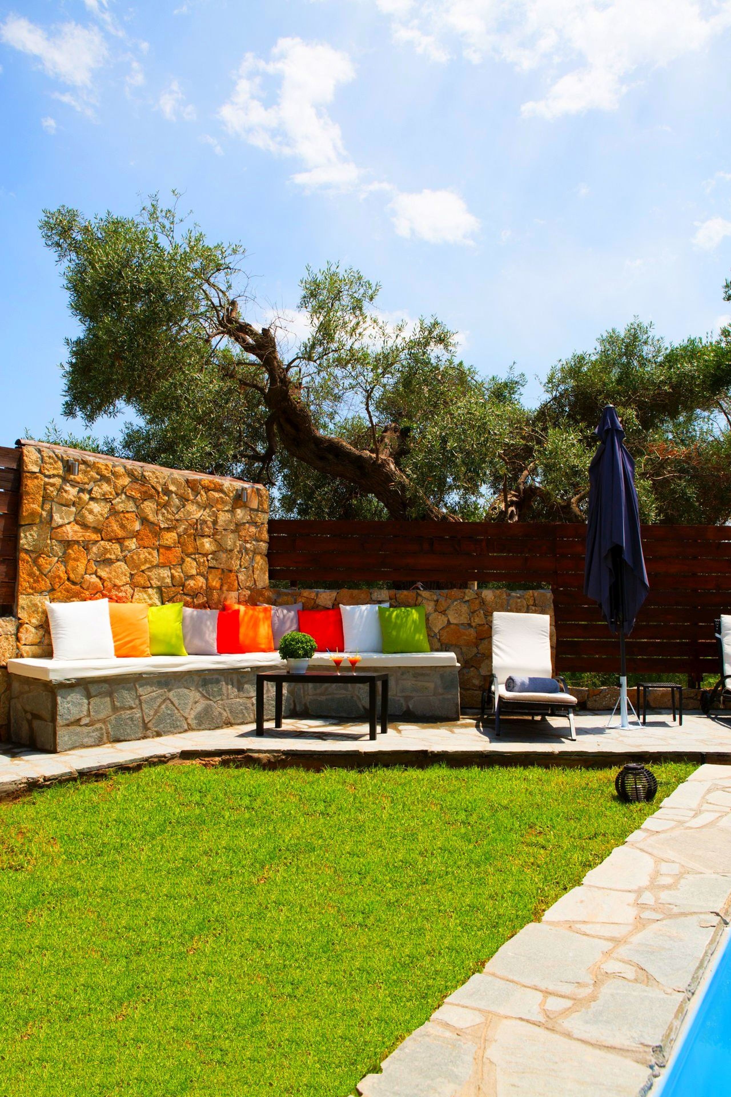 Área de estar com paredes de pedra, almofadas coloridas, uma mesa pequena com bebidas e ambiente à beira da piscina.