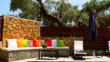 Área de estar com paredes de pedra, almofadas coloridas, uma mesa pequena com bebidas e ambiente à beira da piscina.