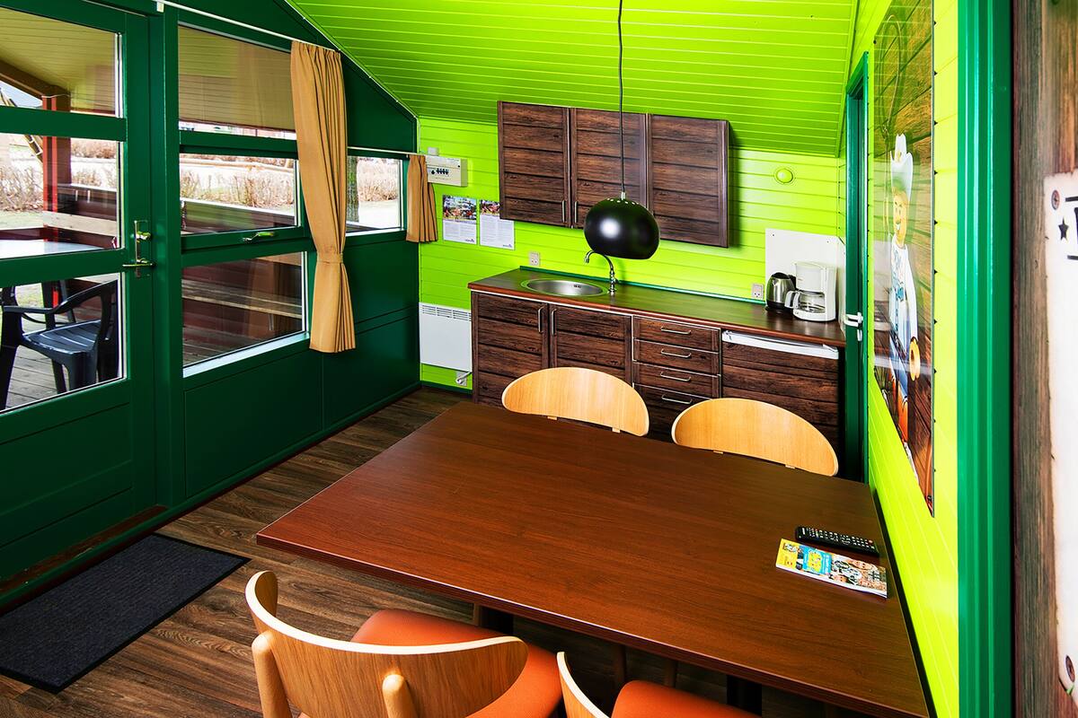 Quarto com mesa de madeira, duas cadeiras, parede verde, janela com cortinas, pia e um cartaz na parede.