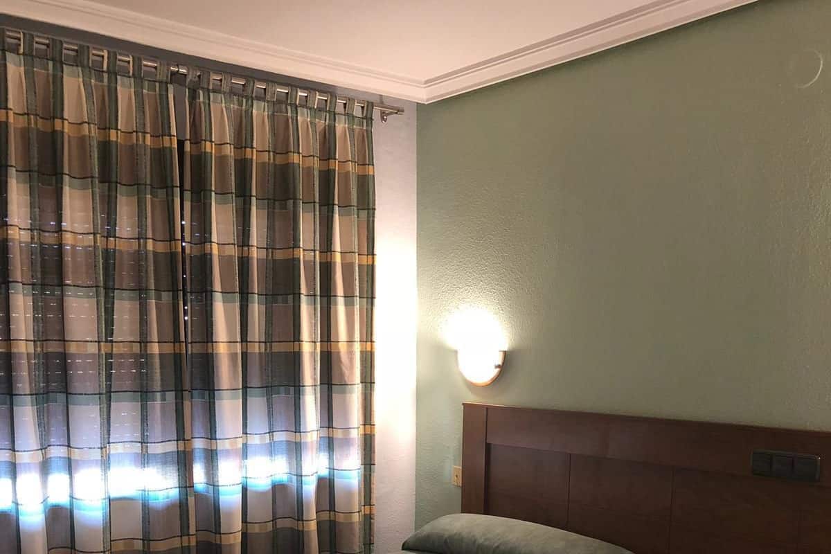 Quarto de hotel com duas camas de solteiro, mesa de cabeceira com telefone, luminária na parede e janela com cortinas xadrez.