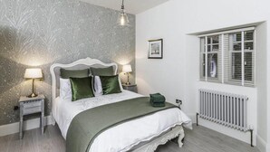 Quarto com cama, duas luminárias de cabeceira, radiador e janela com persianas.