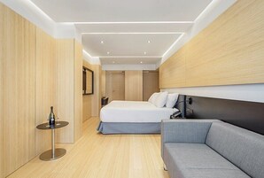 Quarto de hotel moderno com uma cama, um sofá cinzento, uma mesinha redonda com uma garrafa e painéis de madeira nas paredes.