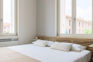 Quarto com cama, duas almofadas, cabeceira de madeira e janelas grandes.