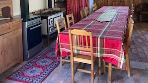 Uma cozinha rústica ao ar livre, com piso de tijolos, mesa e cadeiras de madeira, fogão e churrasqueira.