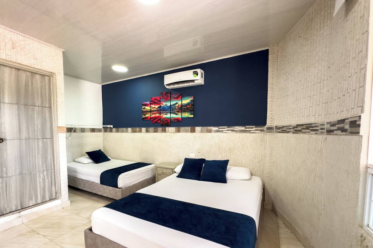 Quarto de hotel com duas camas, uma janela grande, ar condicionado fixado na parede e uma obra de arte colorida acima das camas.