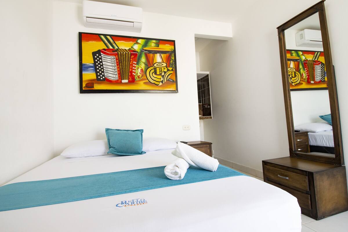 Quarto de hotel com cama, toalha azul, pintura, espelho e cómoda de madeira.