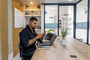 Um homem sentado à mesa de madeira com um laptop, um caderno e um smartphone. Há uma planta em vaso sobre a mesa.