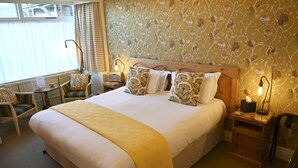 Quarto de hotel com uma cama grande, uma mesa de cabeceira com abajur, uma cadeira, uma mesinha com rádio e uma janela com cortinas.