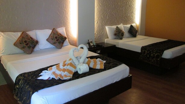 Quarto de hotel com duas camas, cada uma com lençóis brancos e almofadas decorativas. Há uma toalha disposta em formato de coração na cama em primeiro plano.