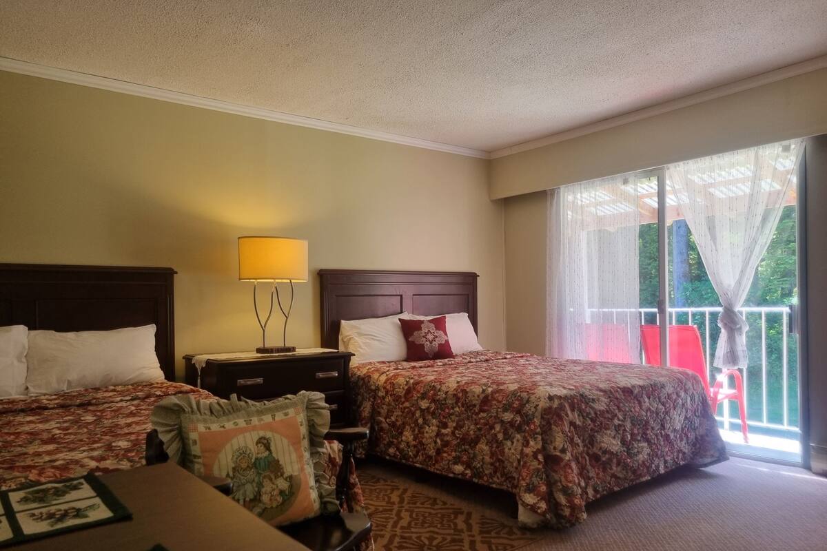 Quarto de hotel com duas camas, uma mesa de cabeceira com abajur, uma pequena mesa com toalha de mesa e vista para o verde pela janela.