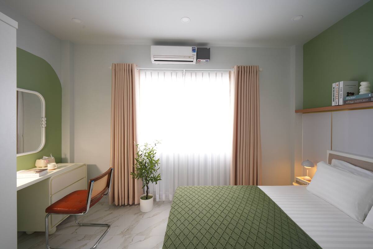 Quarto de hotel com cama, escrivaninha, cadeira, planta em vaso, mesa de cabeceira com abajur e unidade de ar condicionado.