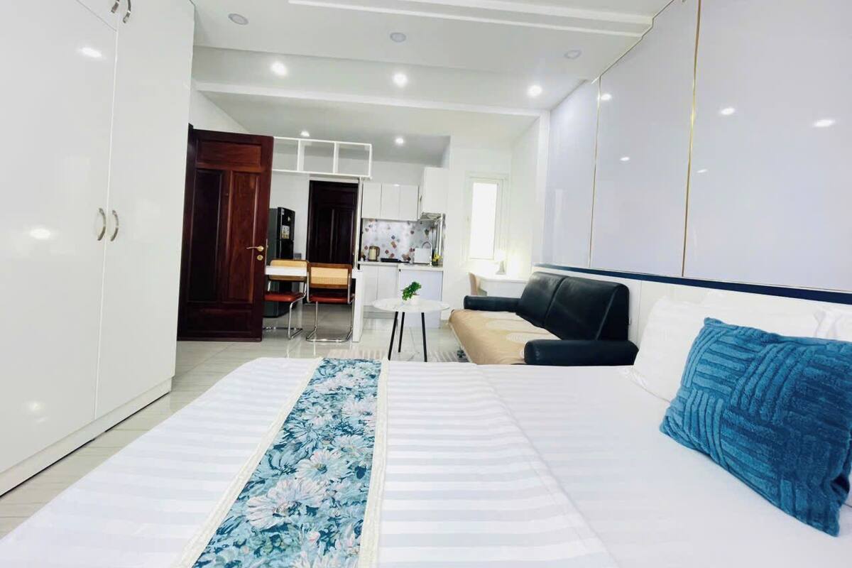 Quarto de hotel moderno com uma cama grande, um pequeno espaço de estar com um sofá, uma cozinha ao fundo e um guarda-roupa.